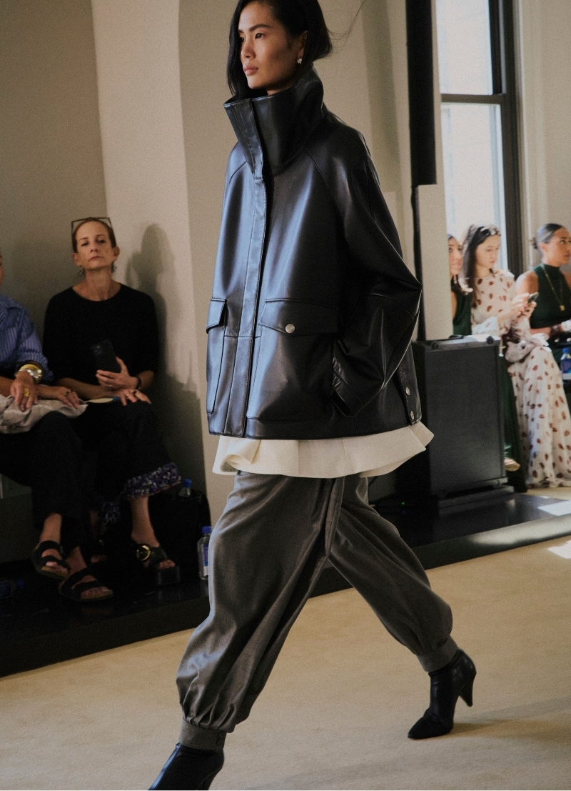 Model in oversized black leather jacket with gray tapered trousers and ankle boots at fashion show