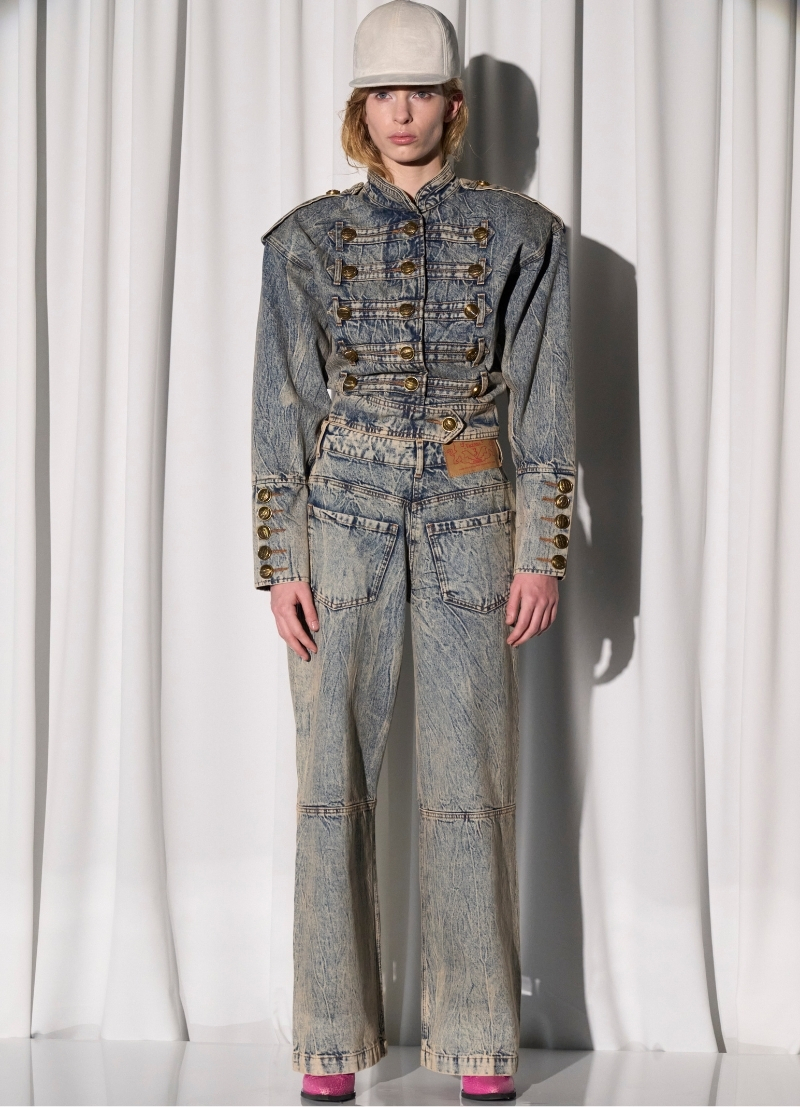 Model in structured denim military jacket and wide-leg jeans with gold buttons against studio backdrop