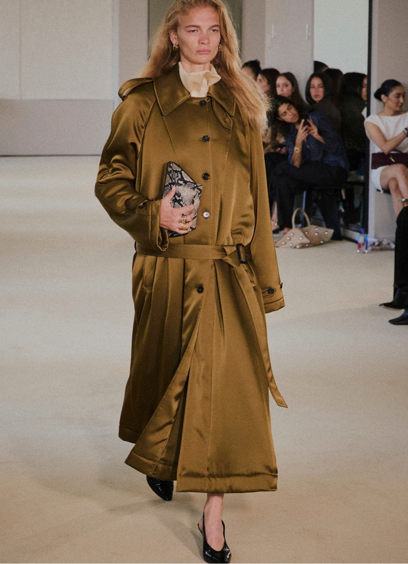 Runway model in an olive satin trench coat with belt and clutch, walking at a fashion show