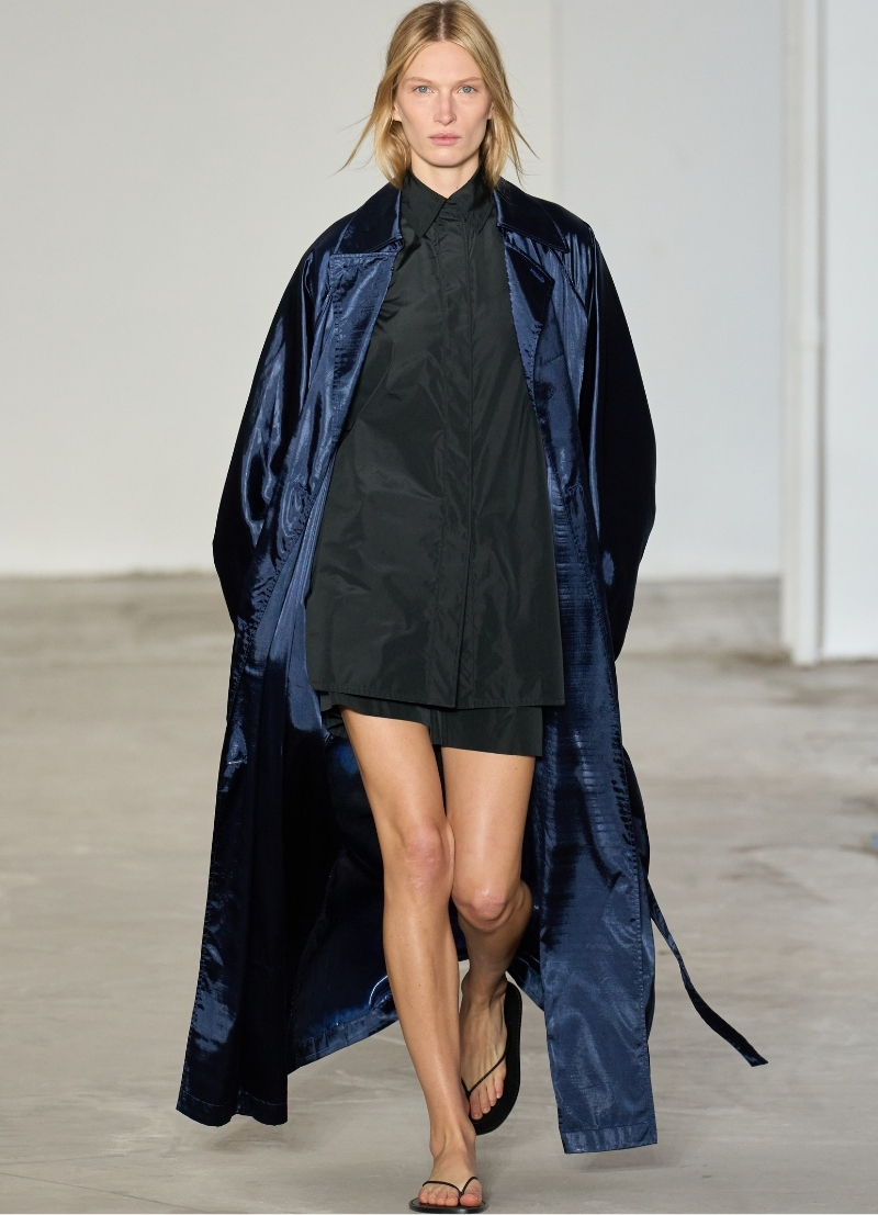 Runway model in a navy satin coat over a black mini dress and thong sandals, on the catwalk