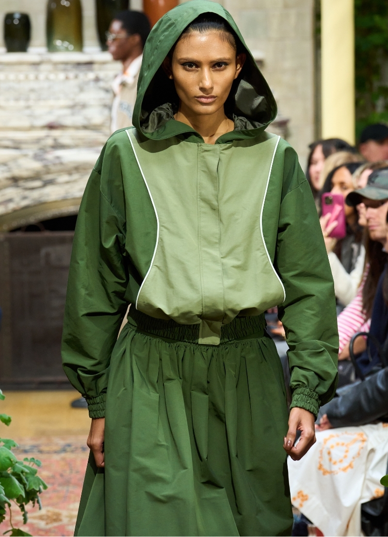 Model in green hooded windbreaker and matching skirt set, styled on indoor runway