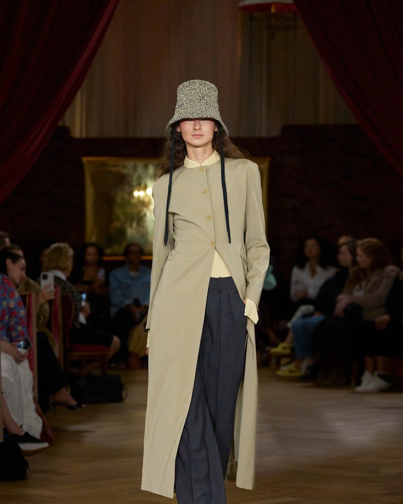 Runway model wearing a beige tailored long coat with wide-leg trousers and a textured bucket hat during a fashion show presentation