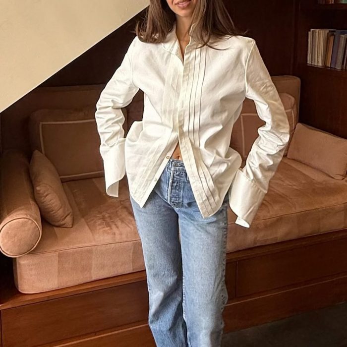 Casual spring outfit featuring a white button-up shirt with pleats, light blue straight-leg jeans, and chic cream flats against a cozy backdrop.
