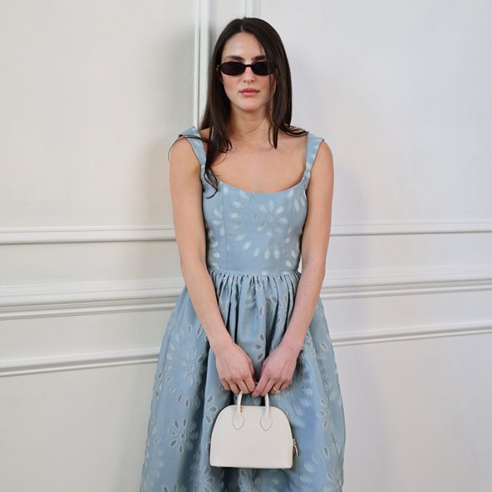 Woman in a light blue floral dress stands against a white wall, wearing sunglasses and holding a small white purse. Calm and stylish atmosphere.