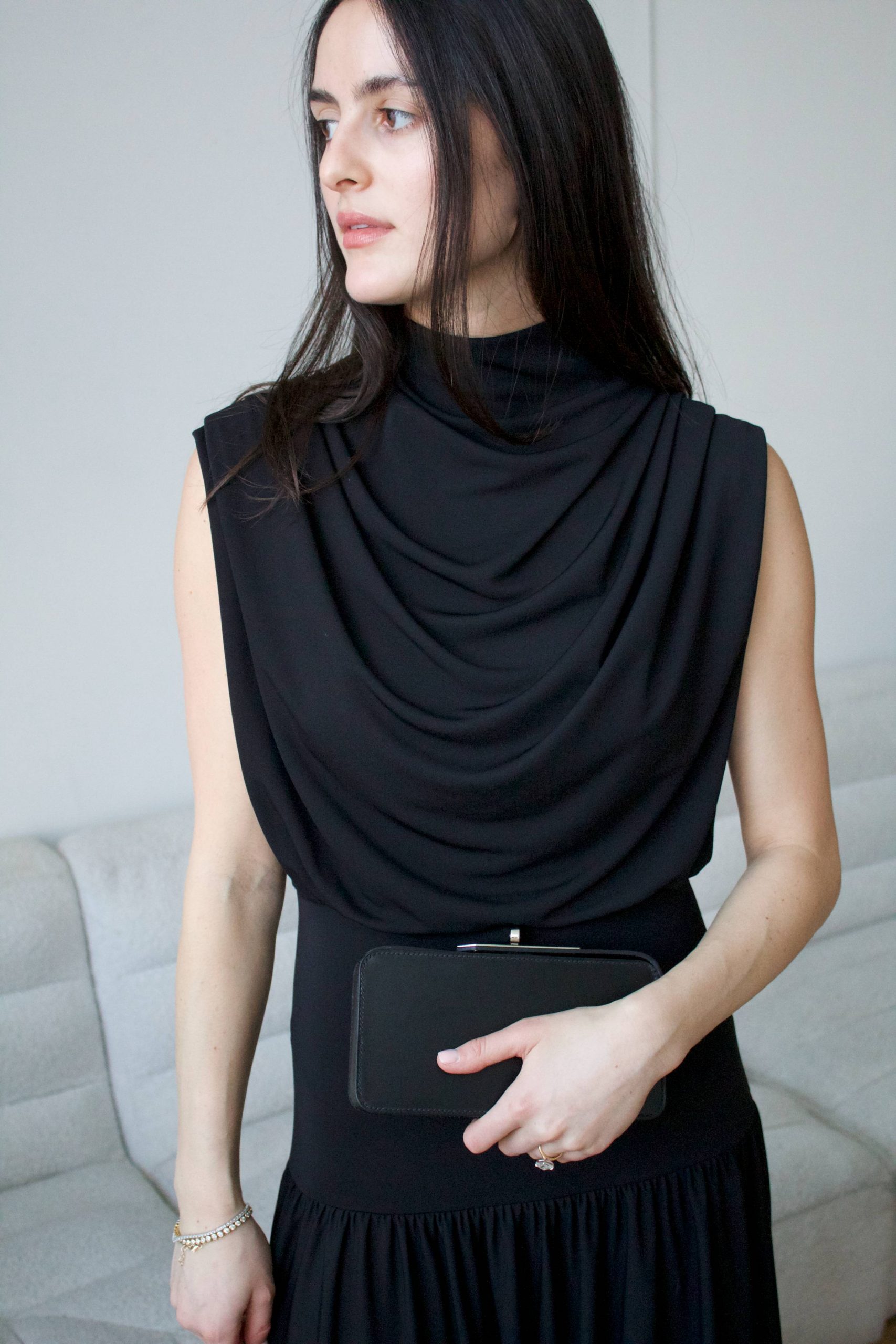 Draped black sleeveless dress with a high cowl neckline and an elegant silhouette%2C styled with a chic clutch and delicate jewelry