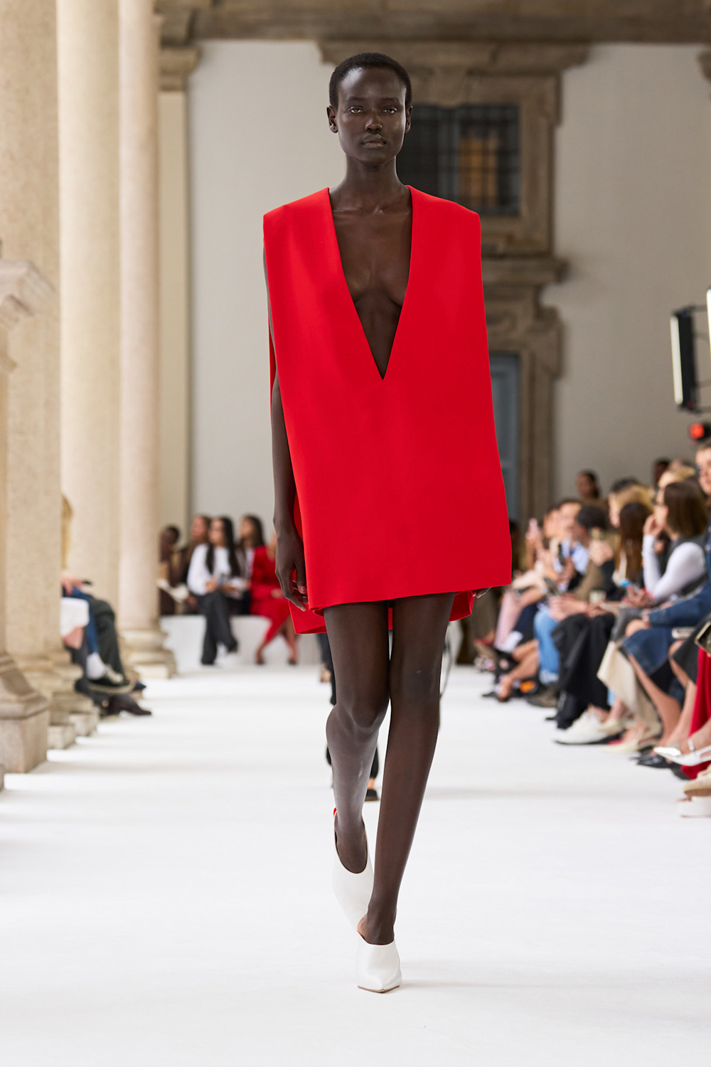 Bold red sleeveless mini dress with a deep V-neck%2C styled with white pointed-toe heels%2C showcasing a striking summer look on the runway