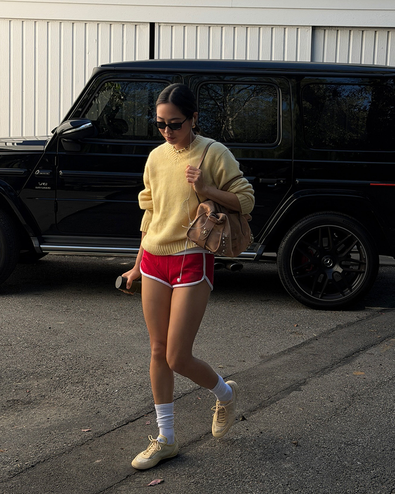 Woman in a cream knit sweater and red athletic shorts with sneakers and a suede shoulder bag on a city street