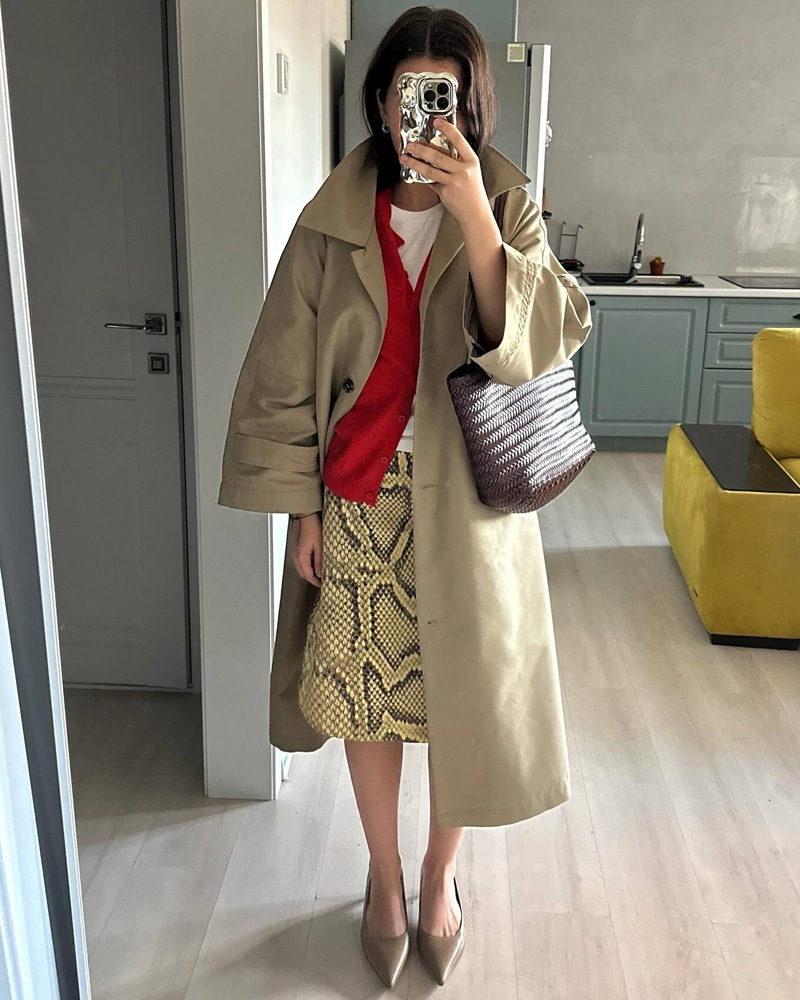 Mirror selfie showcasing beige trench coat layered over red cardigan and snake-print skirt%2C styled with woven tote