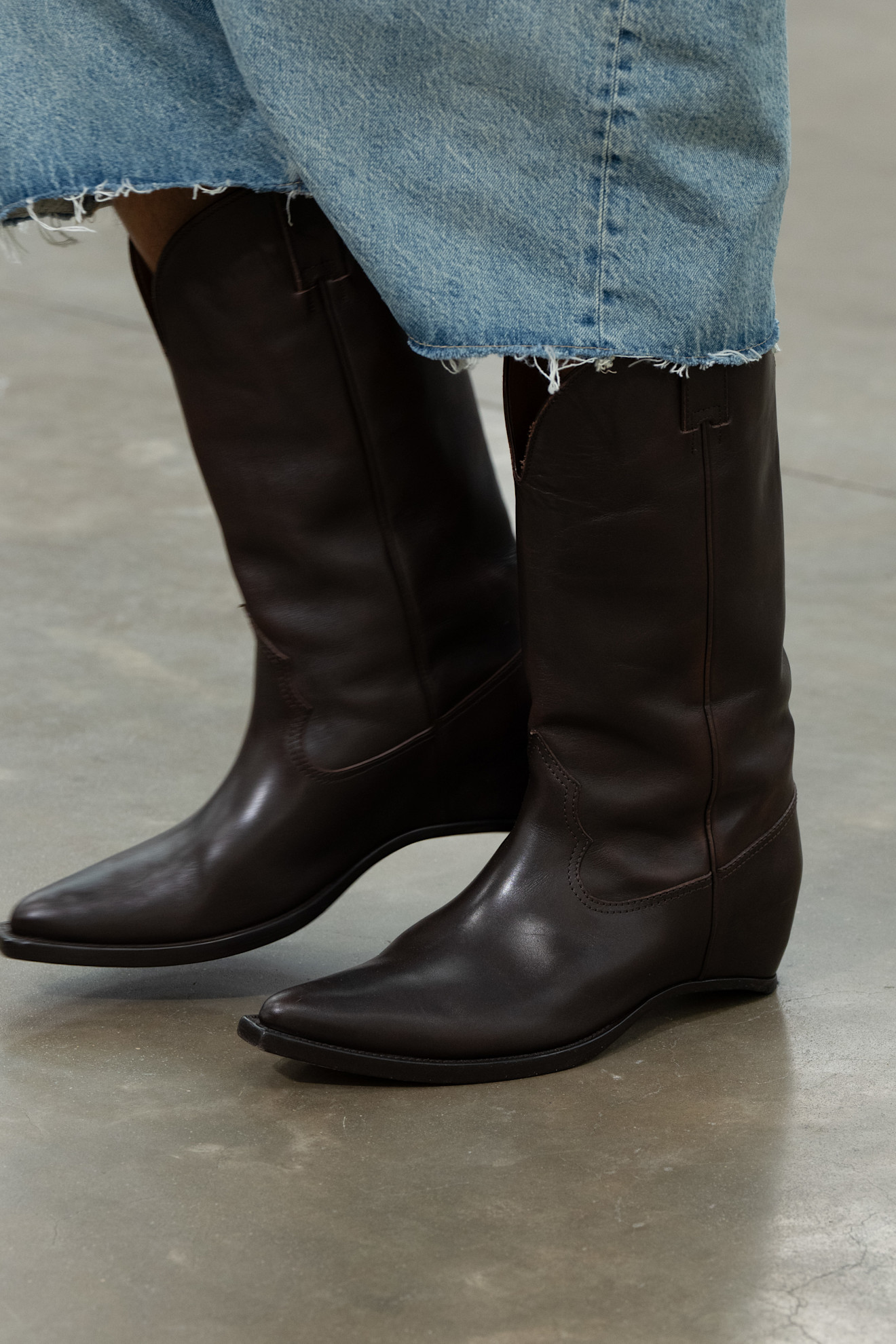 Close-up of a person wearing dark brown leather cowboy boots and blue denim jeans with frayed edges%2C standing on a polished concrete floor.