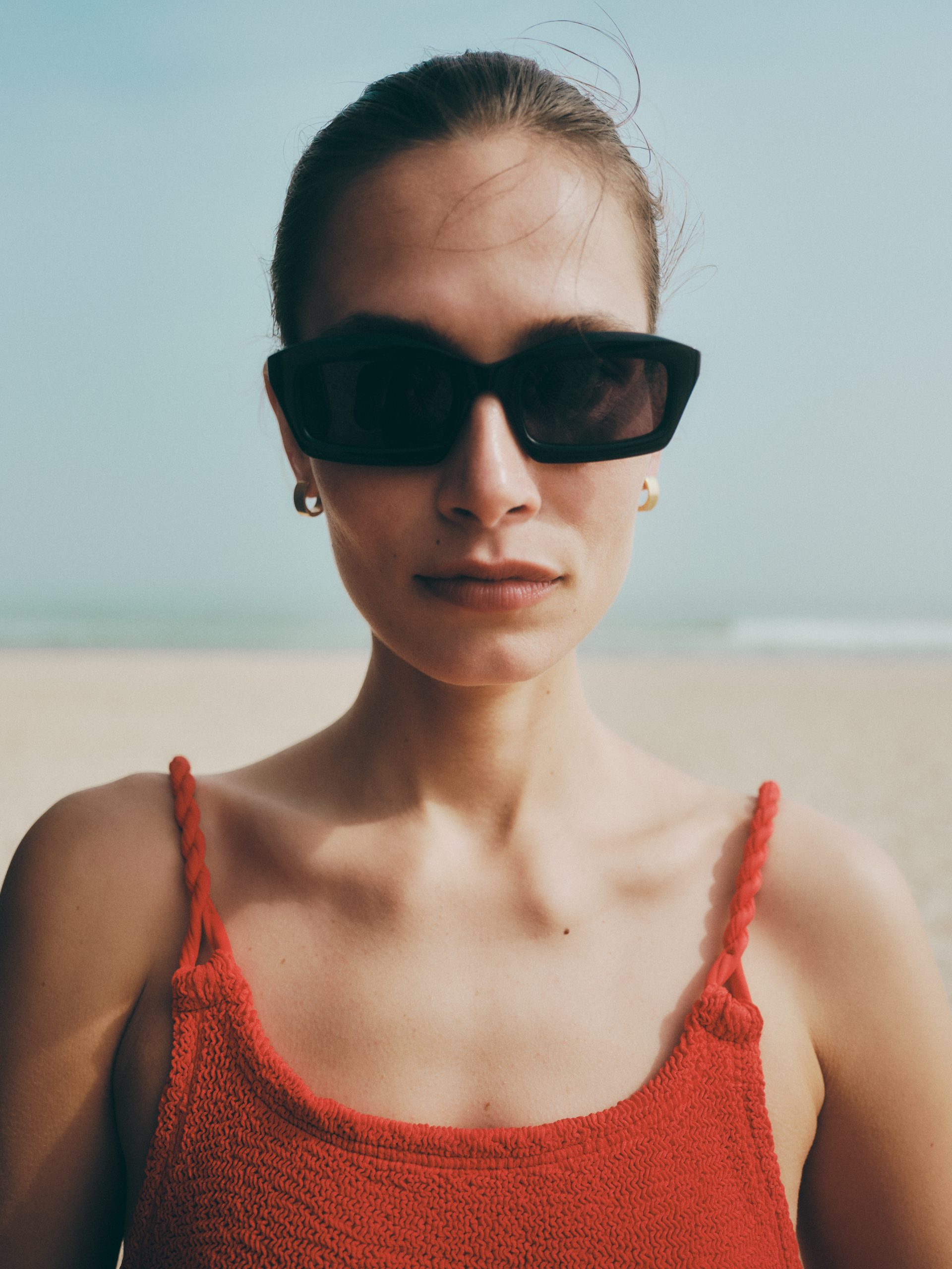Chic seaside look featuring a model in a vibrant red knit tank top with braided straps and stylish oversized black sunglasses%2C set against a beach backdrop