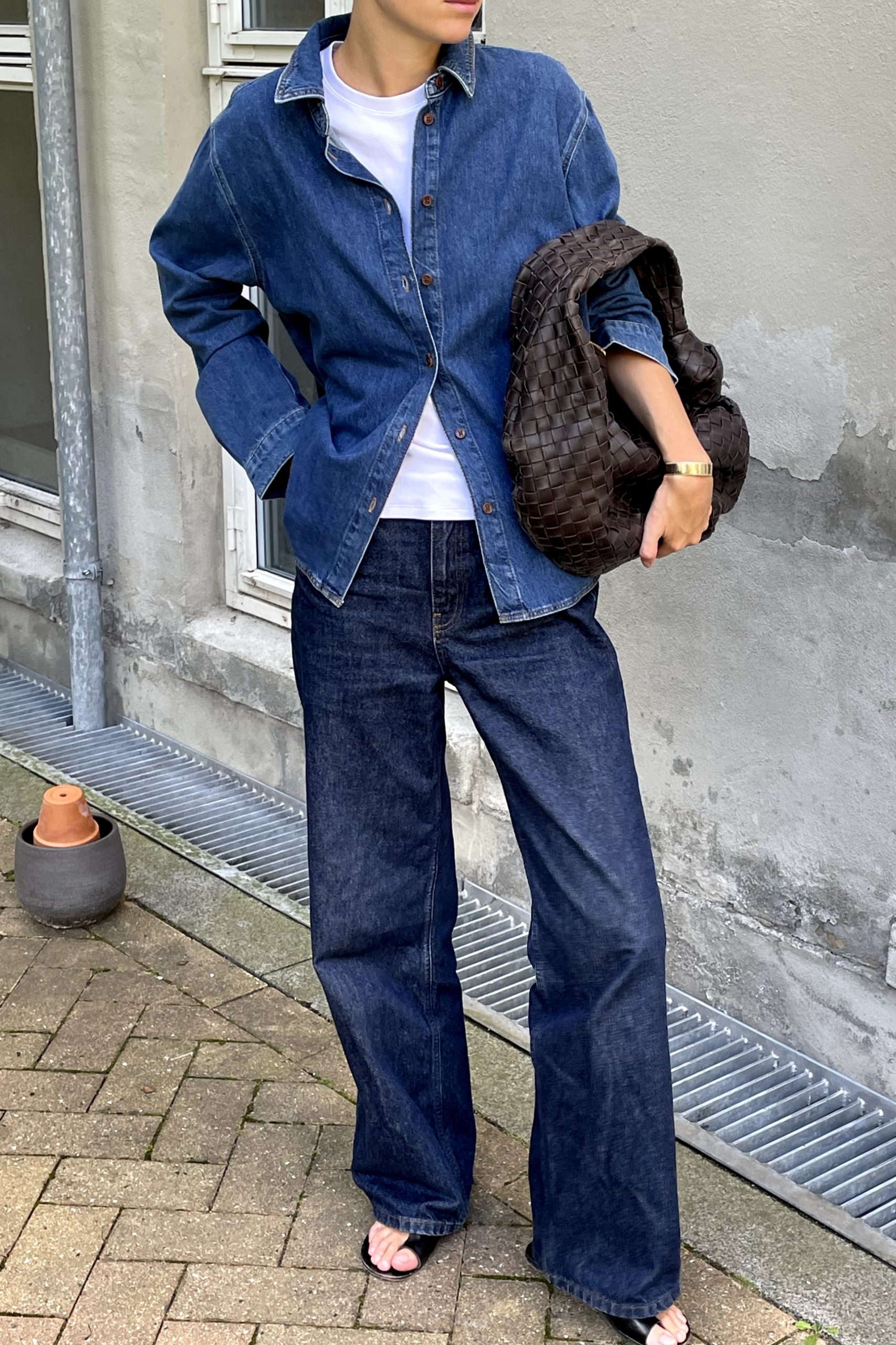 Casual spring outfit featuring a light blue denim shirt layered over a classic white t-shirt%2C paired with wide-leg dark jeans and a woven brown bag