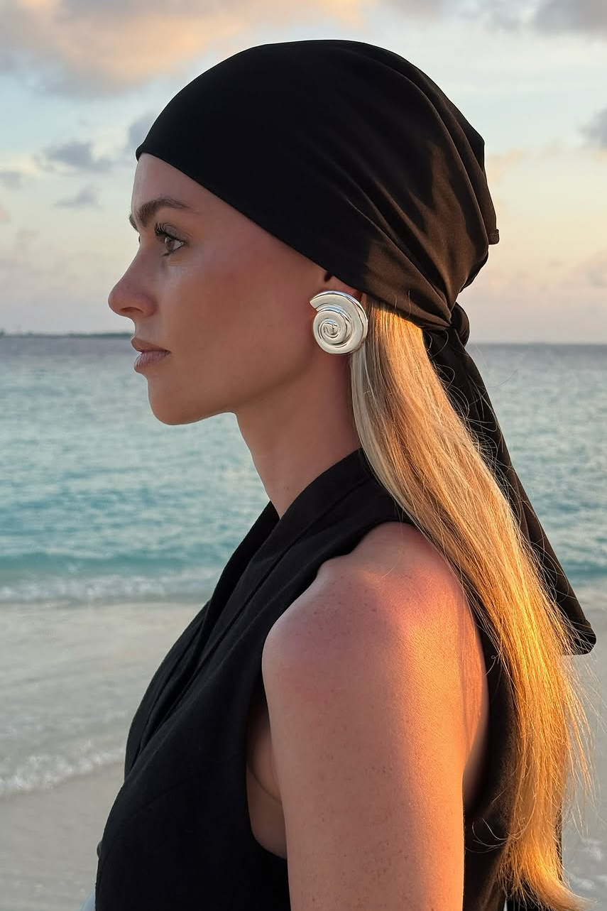 Stylish woman in a black head wrap and sleeveless outfit showcases oversized silver spiral earrings at a beach sunset