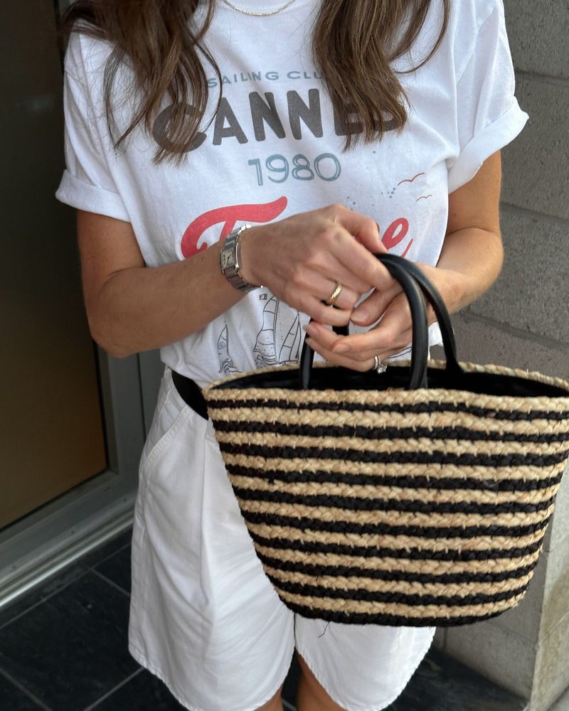 White Cannes 1980 graphic tee with rolled sleeves paired with white high-waisted shorts and a black and natural woven straw tote bag with black handles.