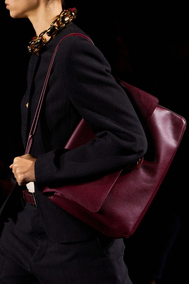 Model wearing a black tailored jacket with a chunky gold chain necklace and carrying a burgundy leather Chanel shoulder bag