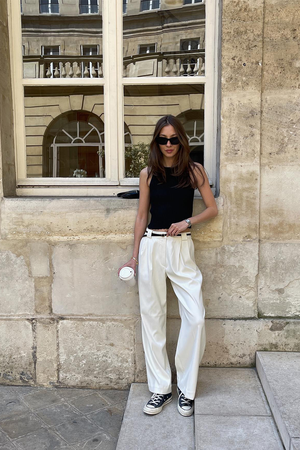 Chic outfit featuring a black tank top%2C tailored white high-waisted trousers%2C and black Converse sneakers%2C styled against a vintage stone backdrop