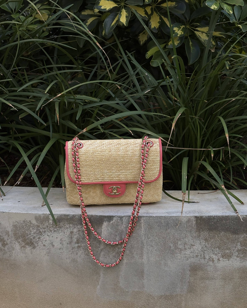 Chanel raffia flap bag with pink leather trim and chain strap%2C styled against lush green foliage for a chic warm-weather vacation look
