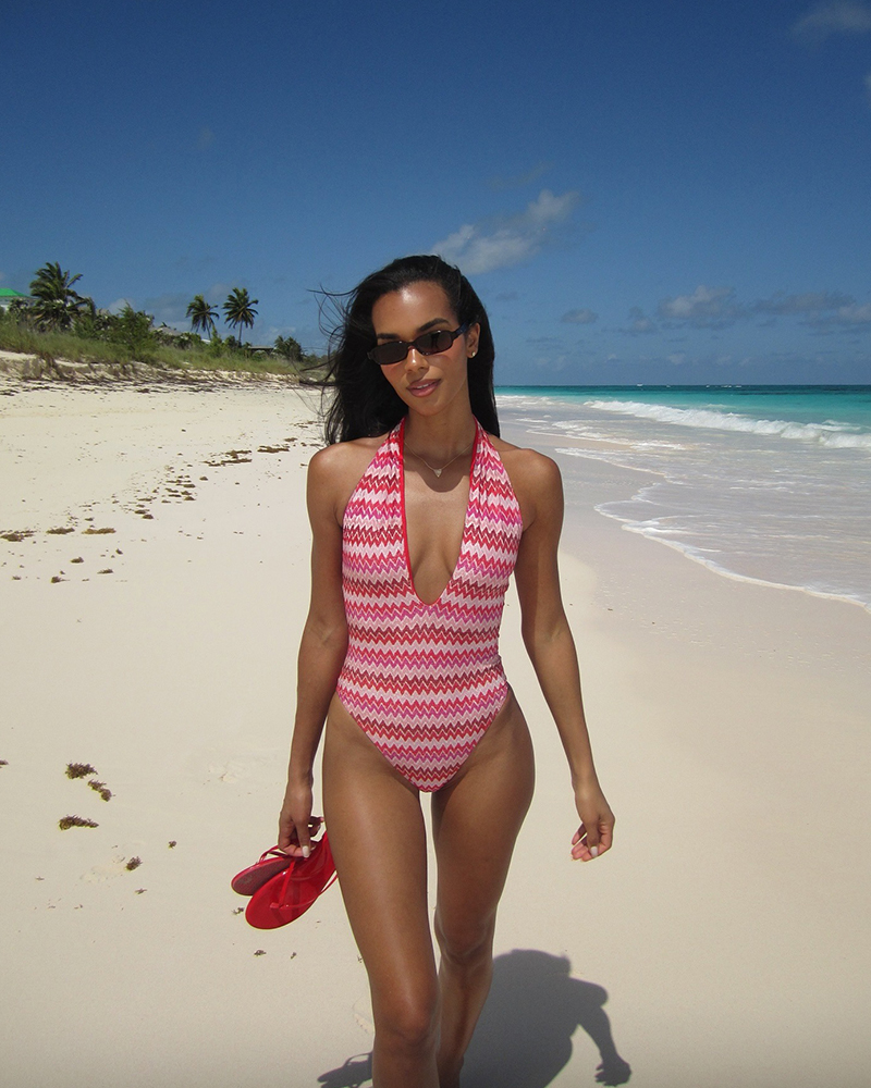 Woman in red zigzag halter one-piece swimsuit with sunglasses and sandals%2C walking along sunny tropical beach shoreline