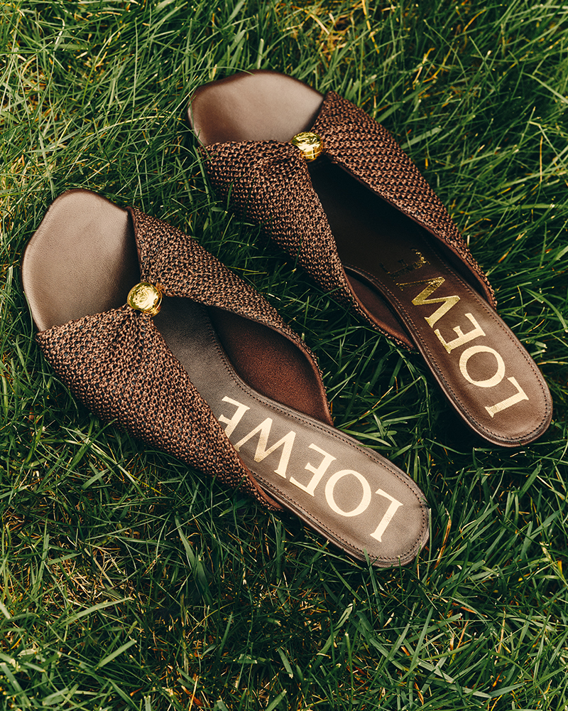 Loewe raffia slide sandals with gold logo detail on lush green grass%2C showcasing a chic summer resort style