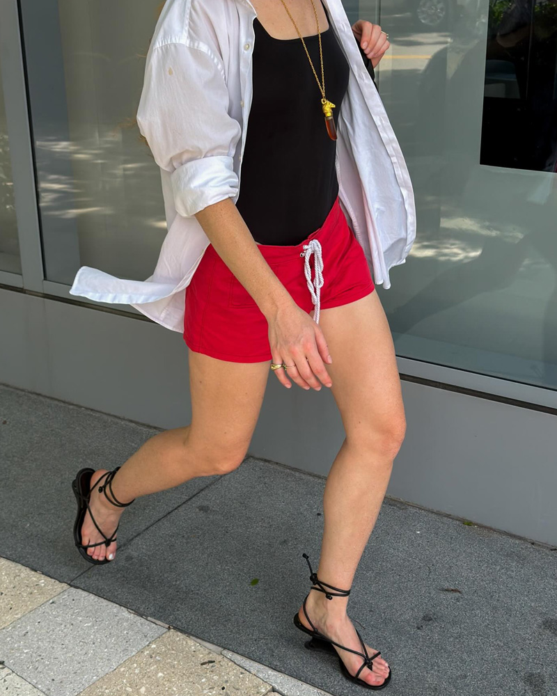 Close-up of summer outfit featuring a white oversized shirt%2C black tank top%2C red drawstring shorts and lace-up sandals on a city sidewalk%0A
