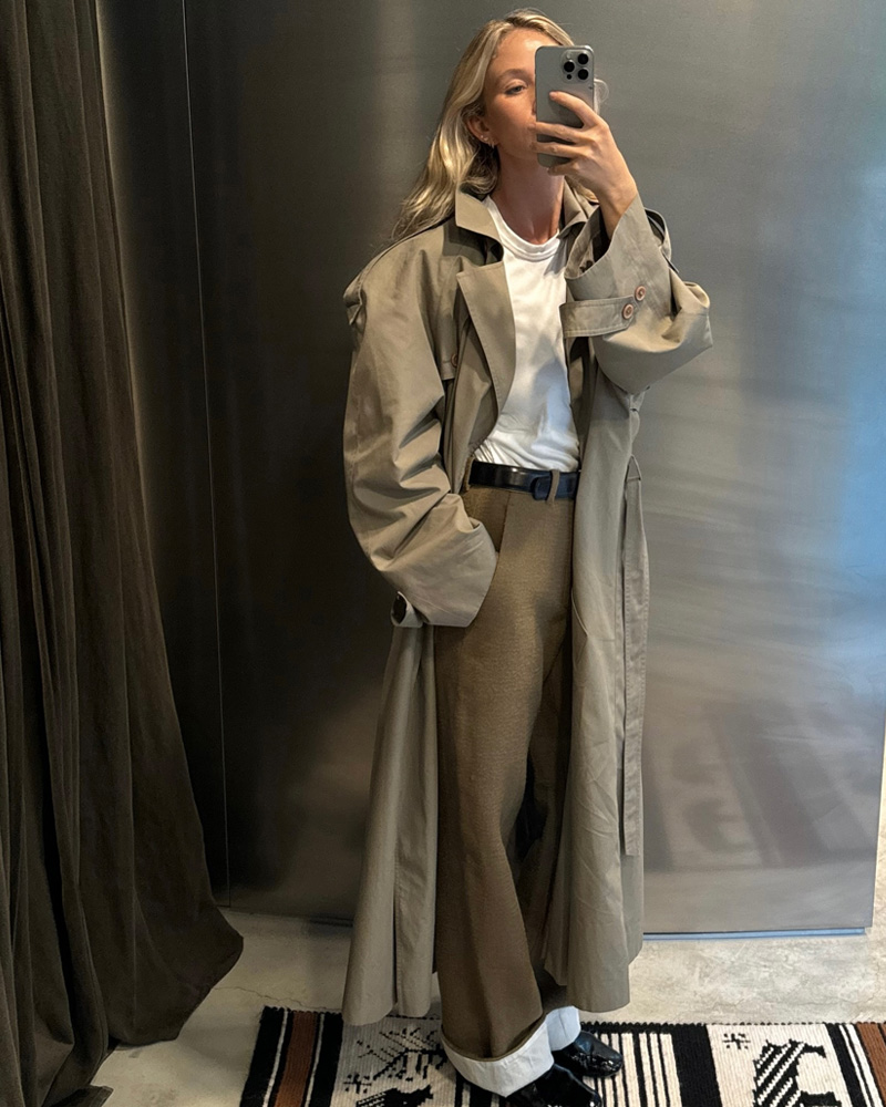 Woman in beige trench coat%2C white tee and brown tailored trousers with loafers taking mirror selfie in minimalist dressing room