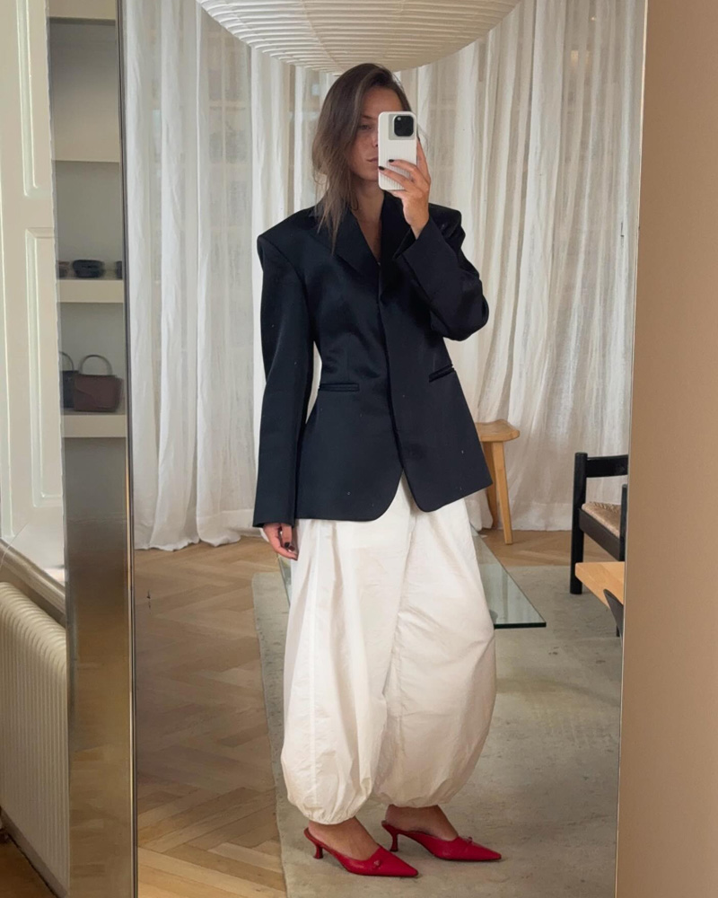 Woman in oversized black blazer and white balloon trousers with red kitten heels taking mirror selfie in minimalist interior