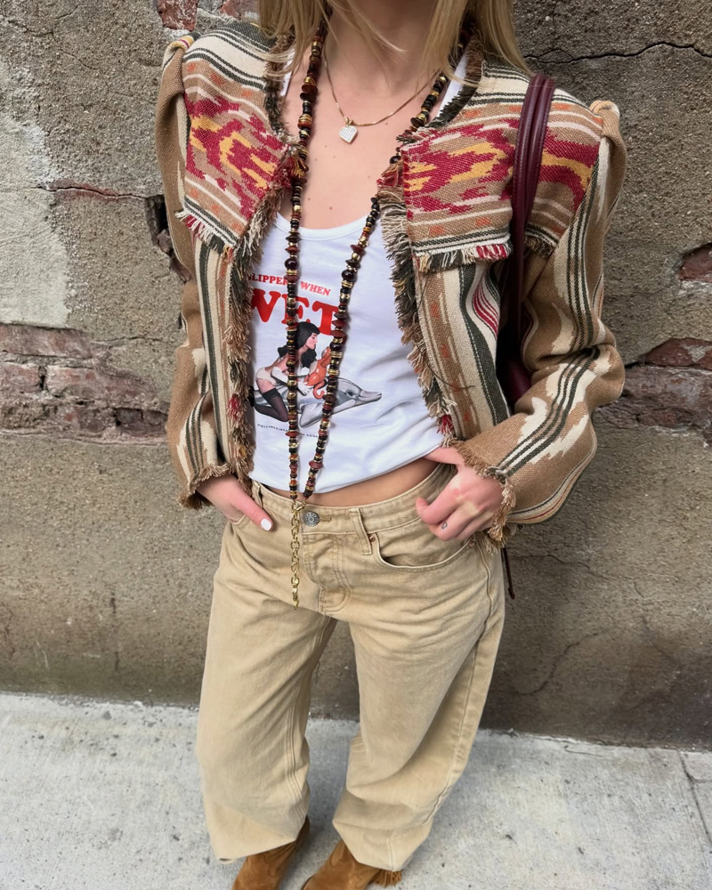 Close-up of boho outfit with fringed patterned jacket%2C graphic tee and beige denim jeans styled against textured stone wall