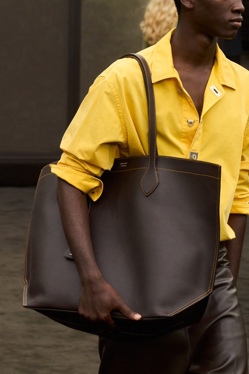 Dark brown leather tote bag with contrast stitching, styled with a mustard yellow shirt and sleek leather trousers