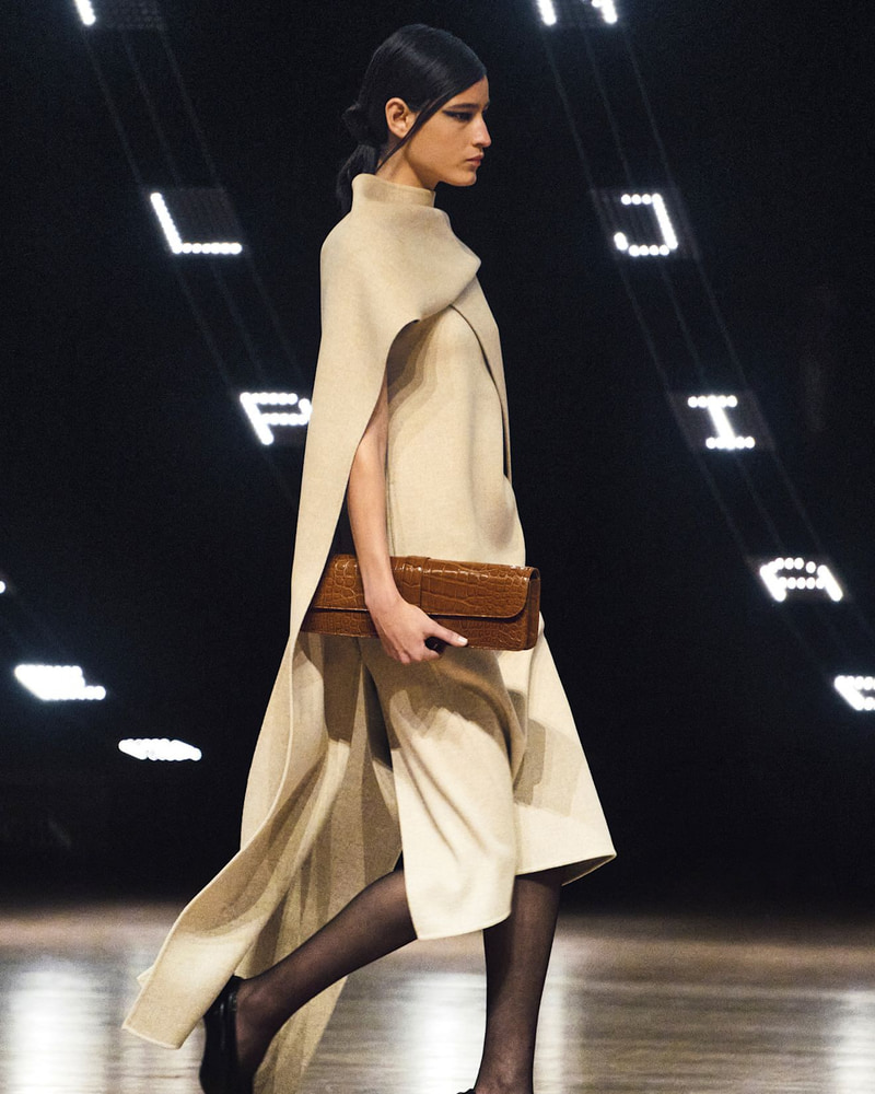 Khaite fall 2026 beige wool cape dress with high collar%2C paired with black sheer tights%2C pointed heels%2C and brown leather clutch