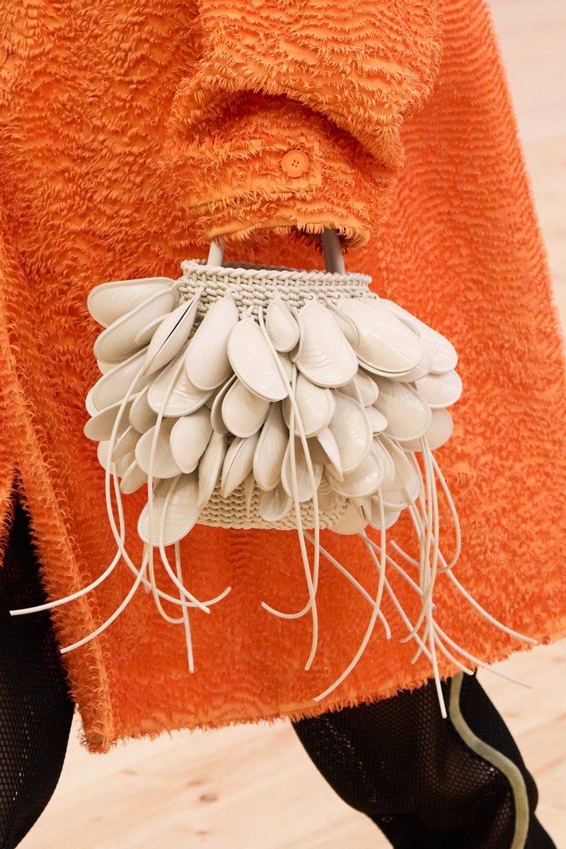 Cream embellished mini handbag with layered petal-like leather details, styled against a textured orange coat for a bold runway look