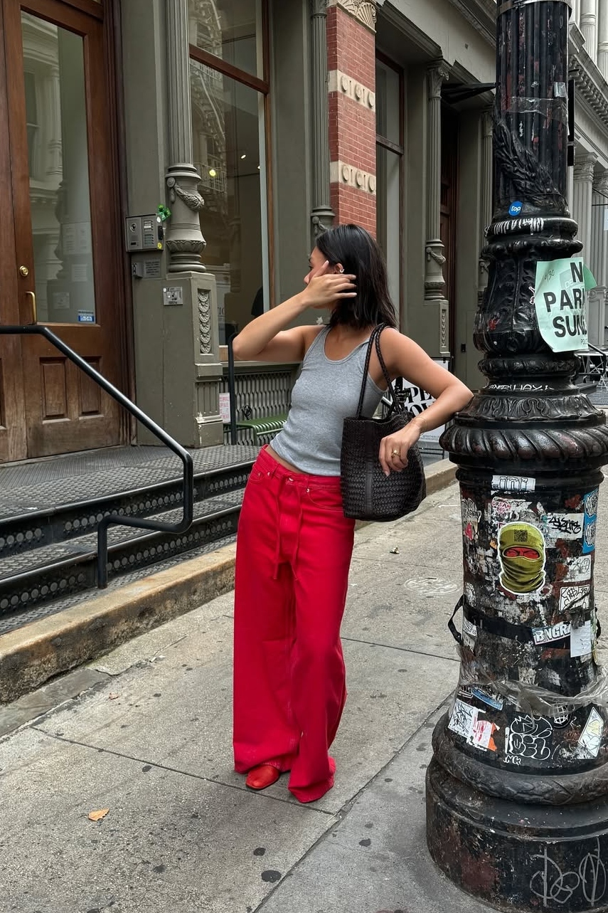 Stylish outfit featuring a gray tank top paired with vibrant red wide-leg pants and matching red heels%2C complemented by a woven black handbag against an urban backdrop