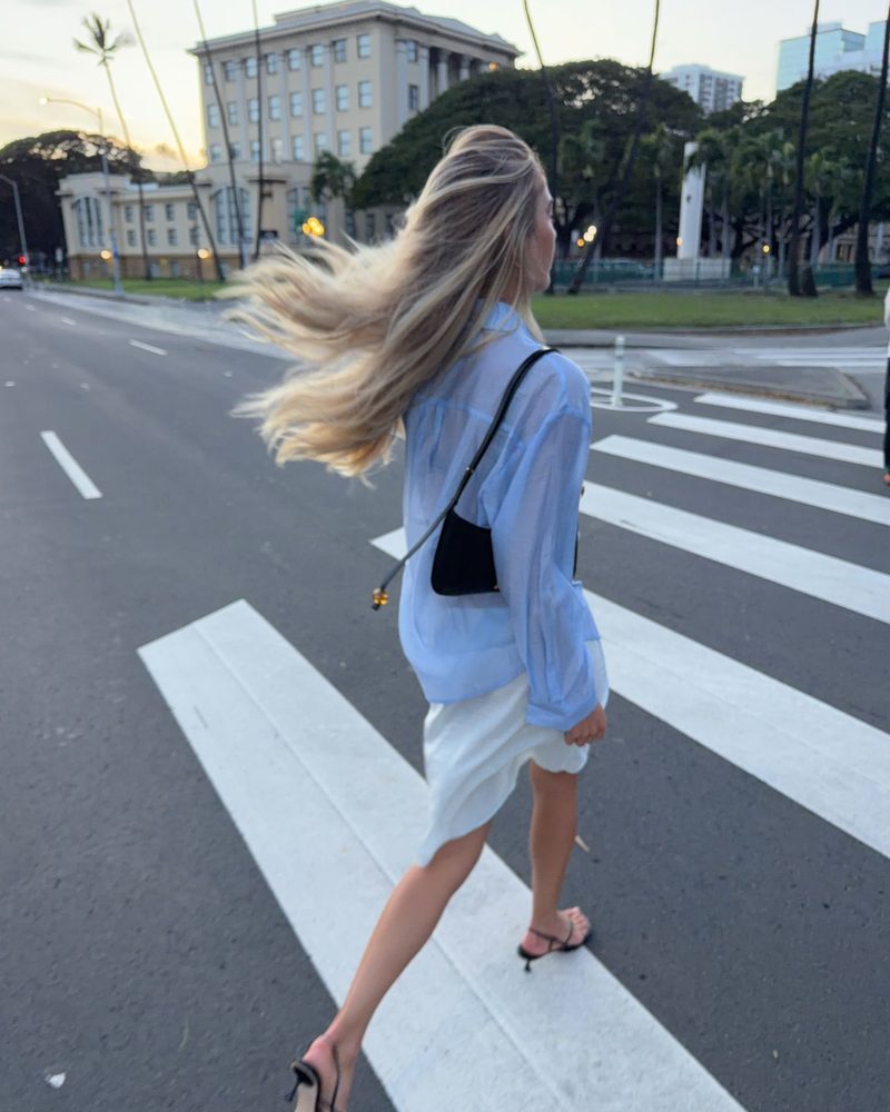 A woman with long blonde hair walks confidently across a city street at dusk%2C wearing a blue shirt and white skirt. Her hair flows%2C conveying motion.