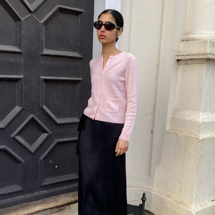 A woman stands outside against a textured black door and white wall, wearing sunglasses, a soft pink cardigan, and a long black skirt, exuding elegance.