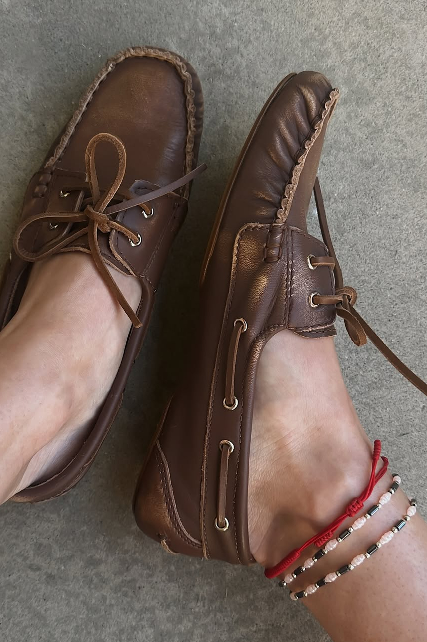 Brown leather moccasin-style loafers with intricate stitching%2C paired with layered anklets featuring beads and a red cord%2C perfect for summer outings