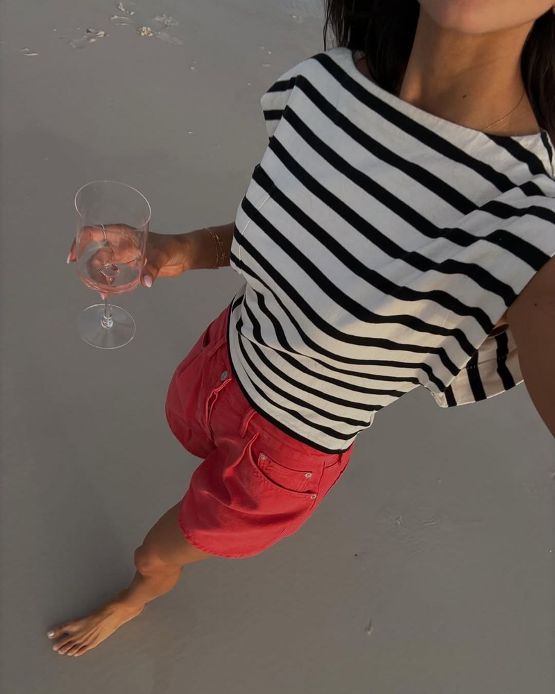Person standing barefoot on sandy ground%2C holding a wine glass%2C wearing a black and white striped top and red shorts.