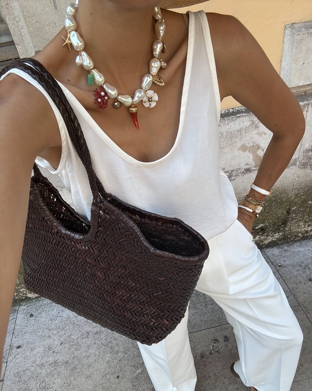Chic layering featuring a statement pearl necklace adorned with colorful charms%2C paired with a relaxed white tank top and woven brown tote bag