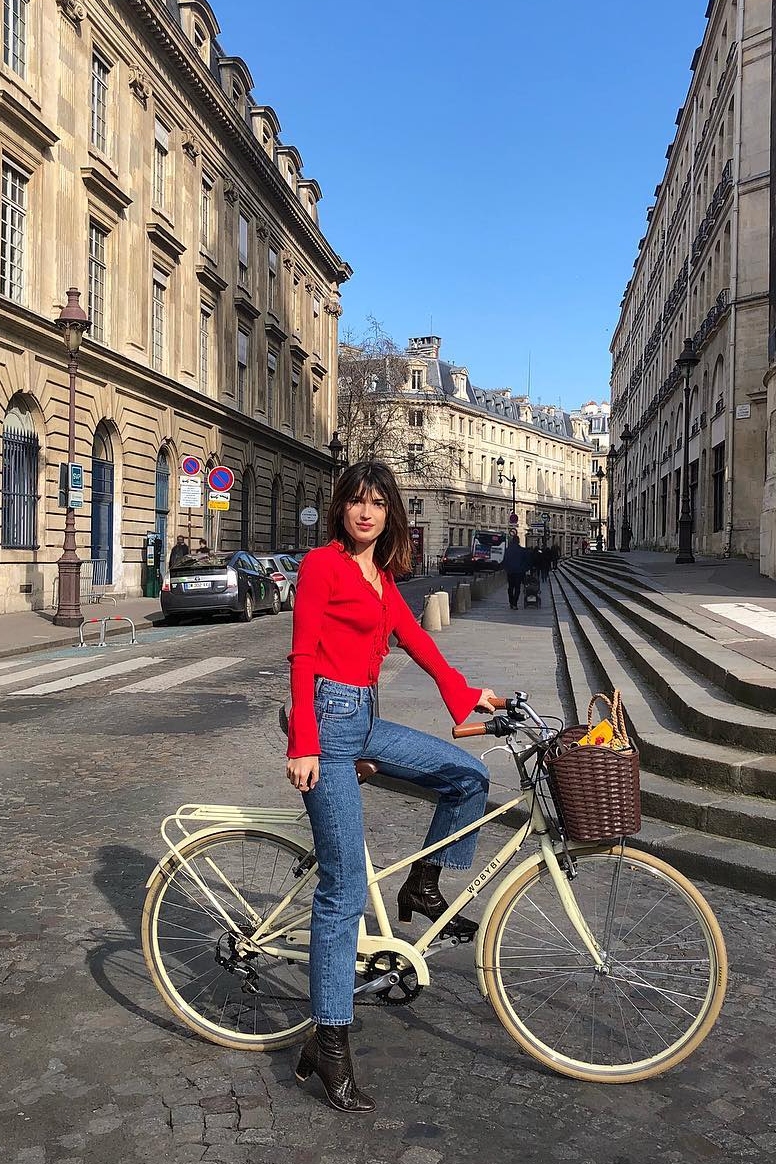 French girl style with a red knit top%2C straight-leg jeans%2C and black ankle boots on a vintage city bike