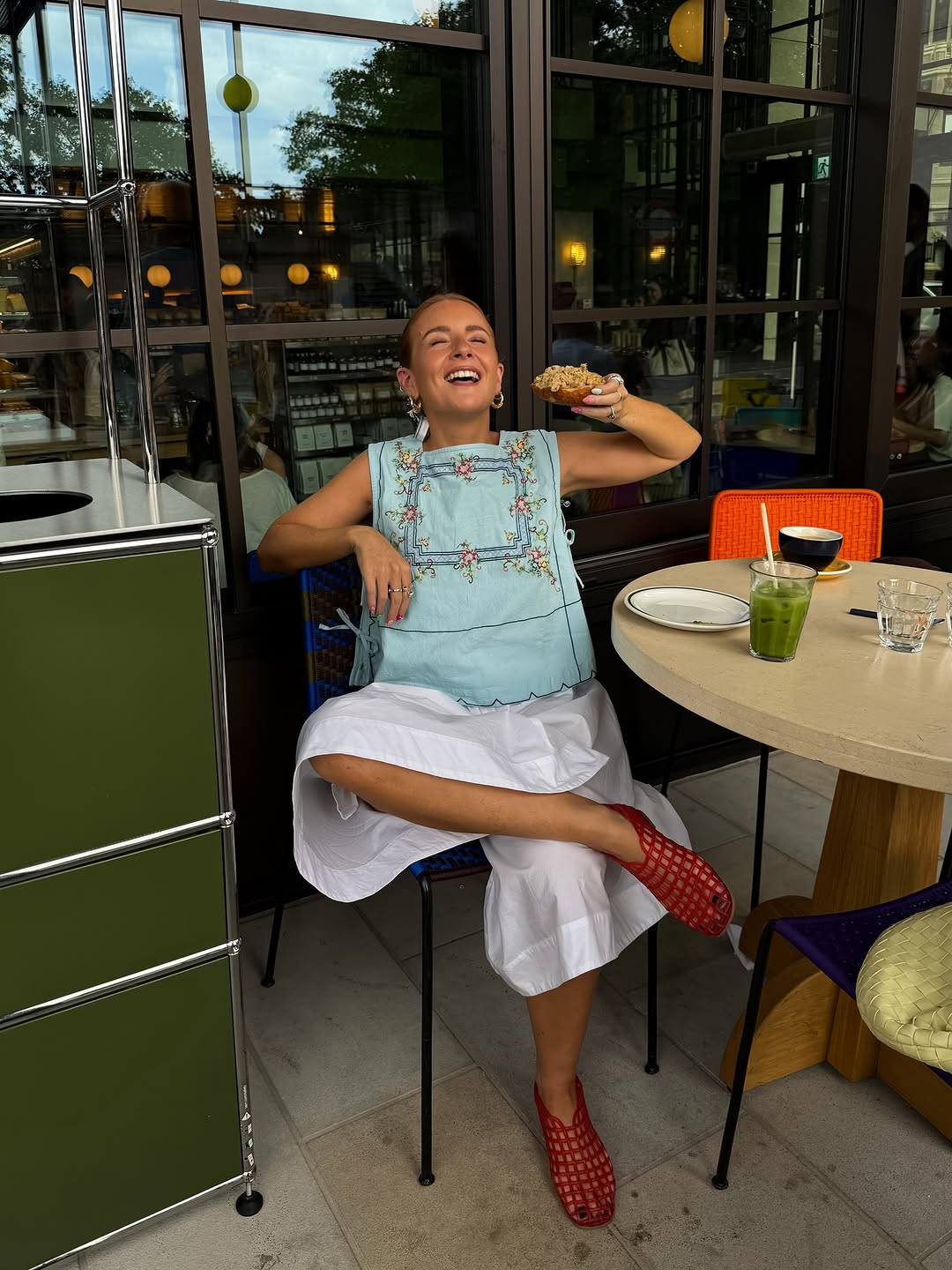 A woman joyfully laughs%2C seated at an outdoor caf%C3%A9 in a blue top and white skirt. She holds a pastry%2C with a green drink on the table beside her.