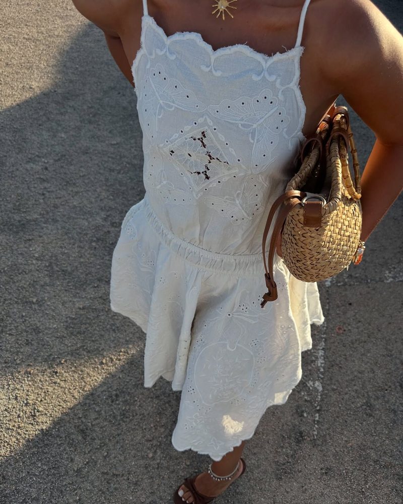 White broderie anglaise summer dress with scalloped edges%2C thin straps%2C paired with a woven straw handbag and brown sandals