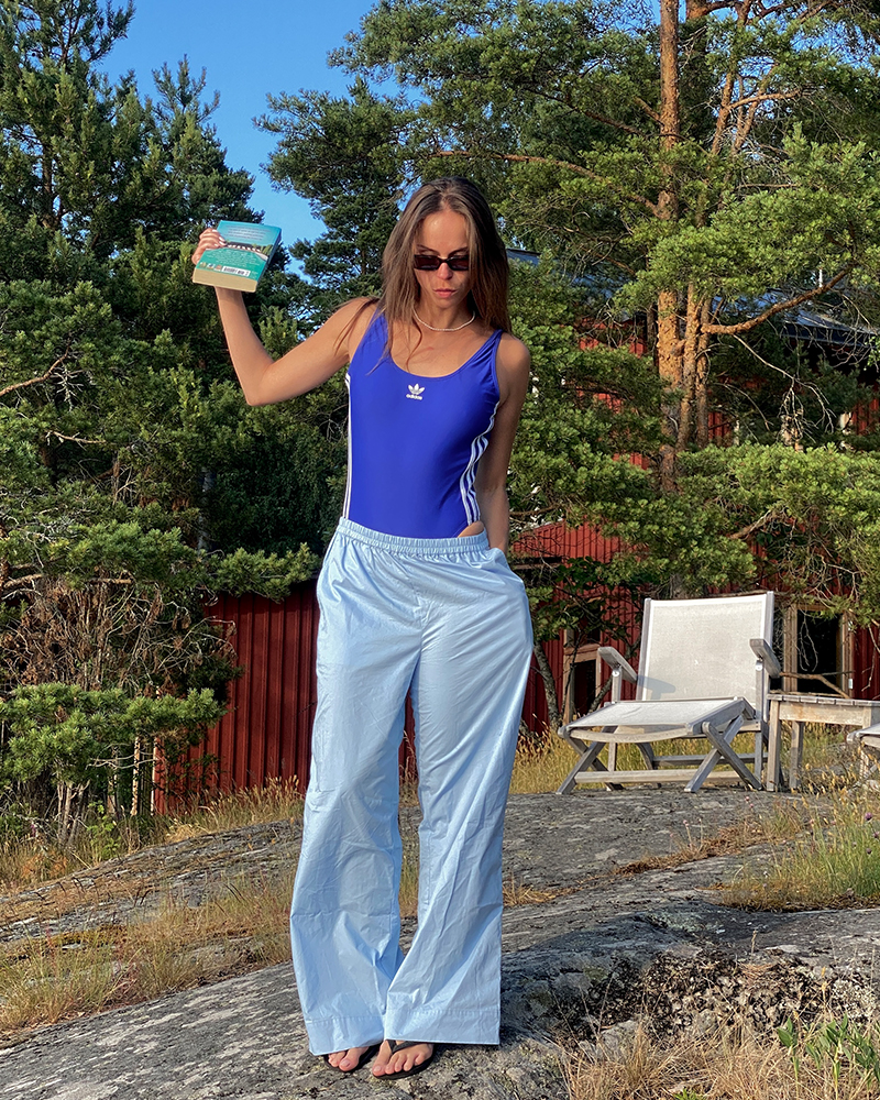 Woman in blue Adidas bodysuit and light blue wide-leg pants with sunglasses%2C standing barefoot in a sunny woodland setting