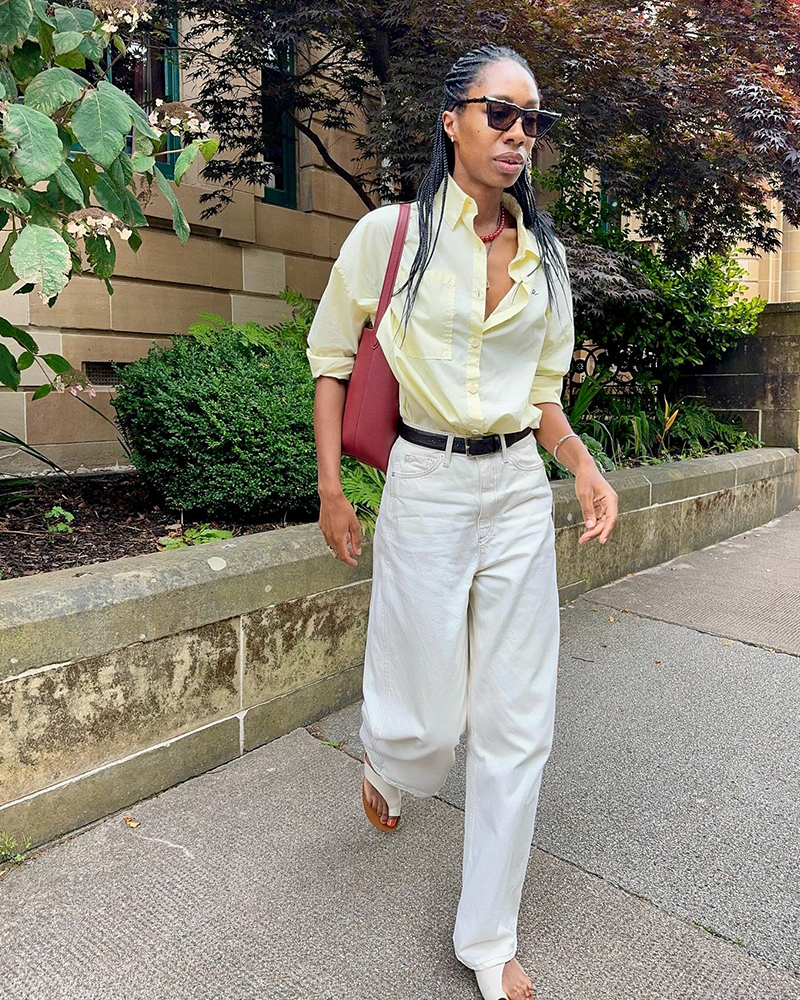Braided hair woman in pale yellow button-down shirt and white wide-leg trousers with red shoulder bag on city sidewalk