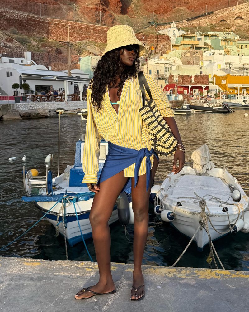 Vacation outfit featuring a yellow striped shirt%2C blue sarong%2C straw bucket hat and woven tote by a scenic seaside harbor