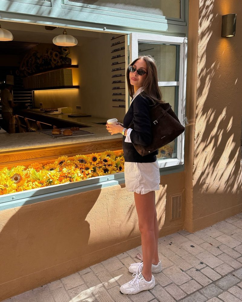 Casual summer outfit featuring a black sweater and white shorts%2C paired with white sneakers and a brown shoulder bag against a sunny backdrop