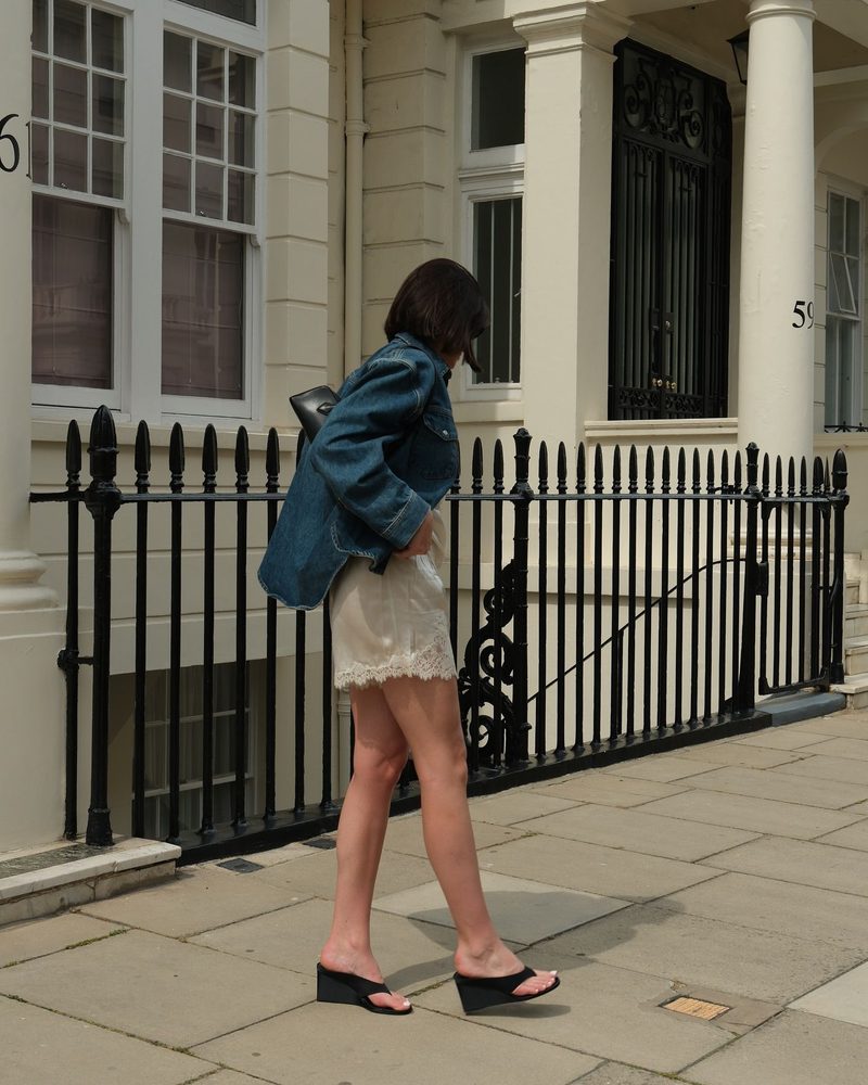 Chic summer outfit features a denim jacket paired with a lace-trimmed white mini skirt and black wedge sandals%2C set against an elegant urban backdrop