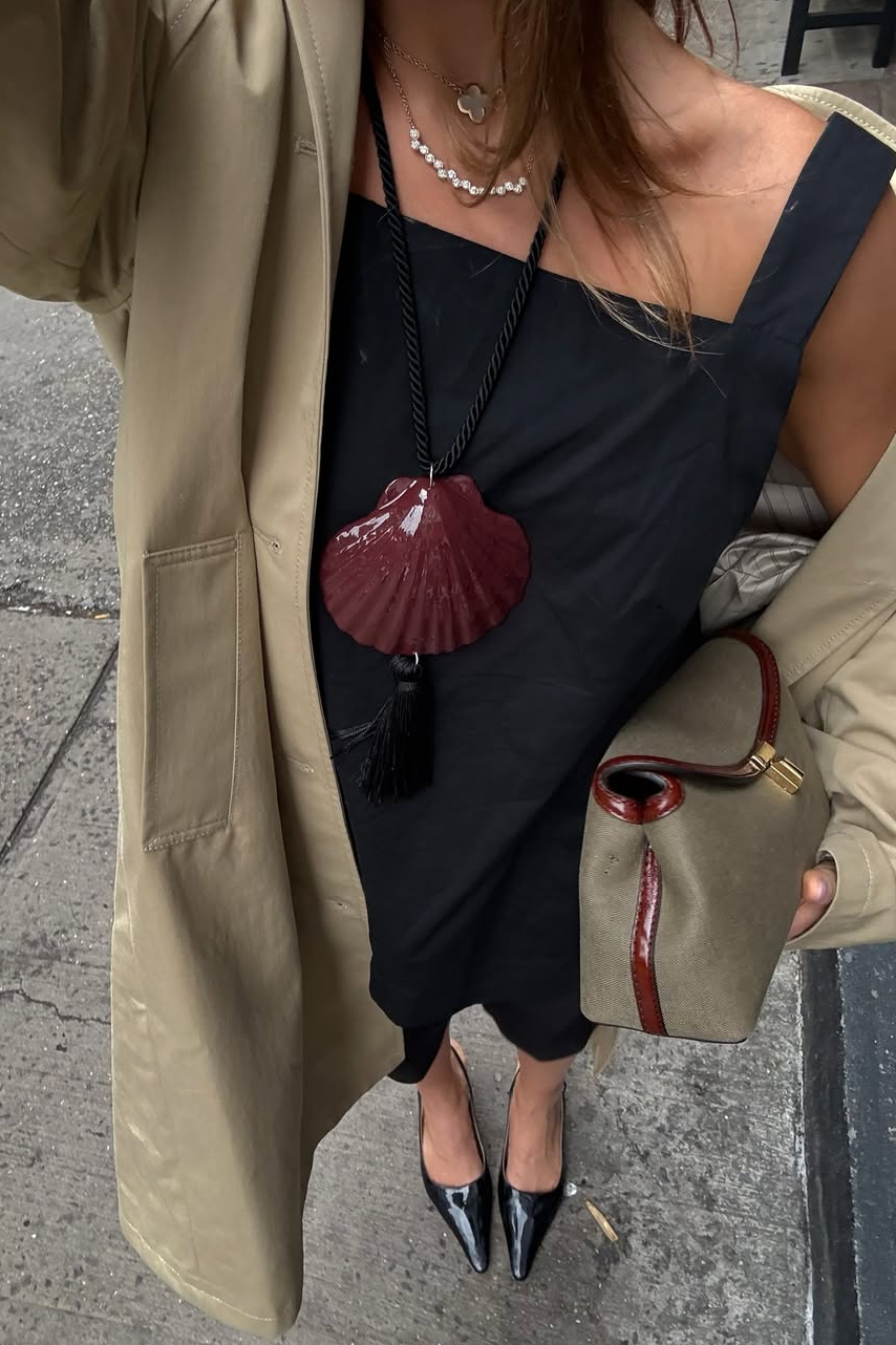 Chic monochrome outfit featuring a black slip dress layered with an oversized beige trench coat%2C accessorized by a statement red shell cord necklace with a tassel