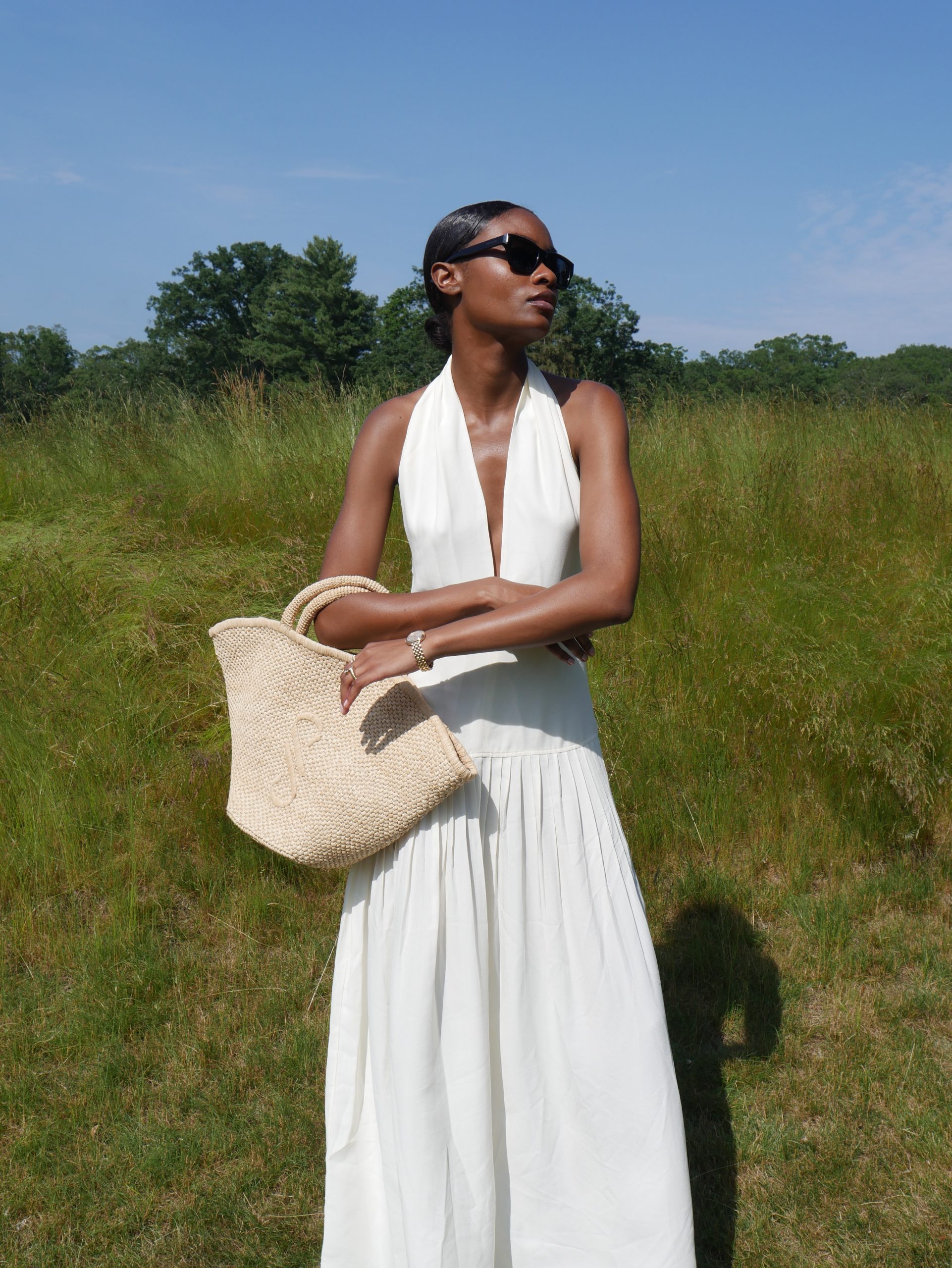 Elegant Jacquemus halter neck maxi dress in white%2C paired with a woven straw bag and chic black sunglasses%2C set against a clear blue sky