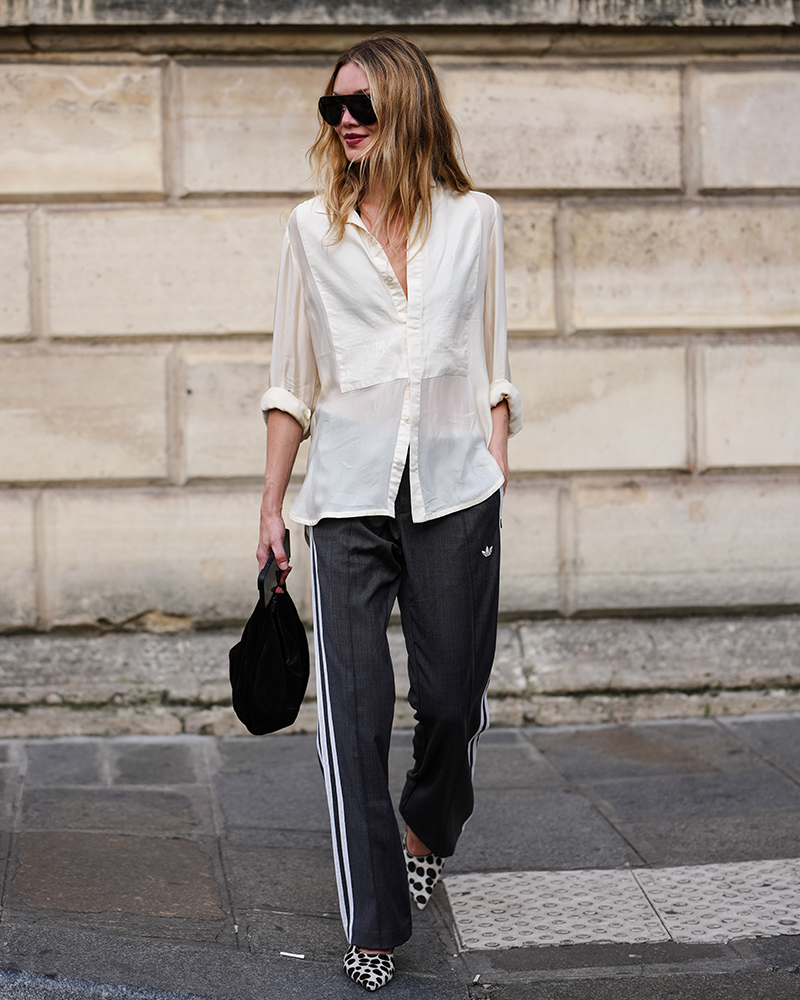 Chic woman in a sheer white button-up shirt paired with gray Adidas track pants and stylish leopard-print heels%2C accessorized with oversized sunglasses and a black bag
