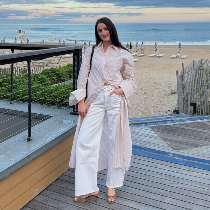 A woman poses on a wooden deck by the beach at sunset, wearing a striped shirt and white pants. The ocean and a cloudy sky form the backdrop.