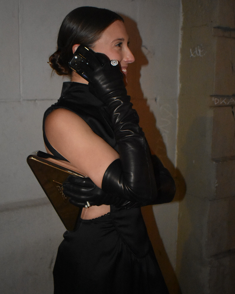 Evening look featuring a black satin gown with opera-length leather gloves%2C styled with a gold clutch and statement ring in a dim city setting