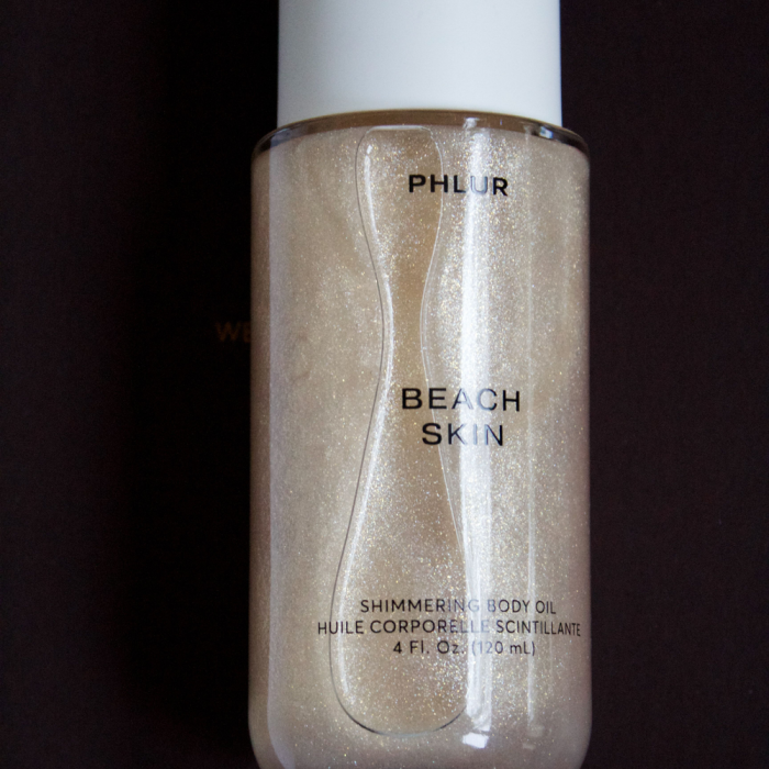 Clear glass bottle of shimmering body oil with a white cap, labeled "Phlur Beach Skin." The golden liquid inside radiates a subtle sparkle.