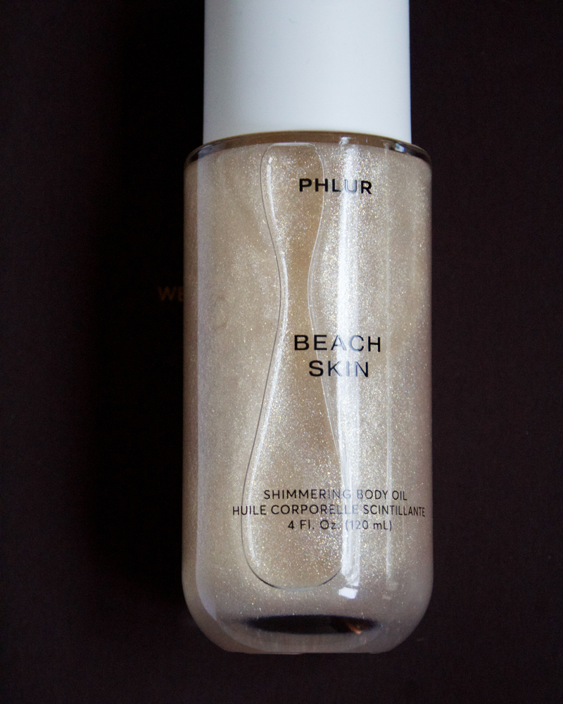 Clear glass bottle of shimmering body oil with a white cap%2C labeled %22Phlur Beach Skin.%22 The golden liquid inside radiates a subtle sparkle.